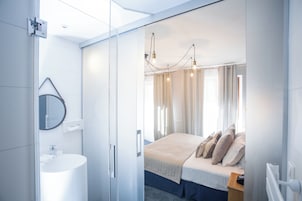Quarto de hotel moderno com divisória de vidro separando o banheiro e o quarto. O banheiro possui uma pia branca e um espelho. O quarto tem uma cama com cobertor e almofadas bege, uma mesa de cabeceira de madeira e um lustre.