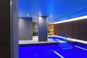 Uma área interna de piscina moderna, com piscina iluminada em azul, parede de madeira e um espaço de estar ao fundo.