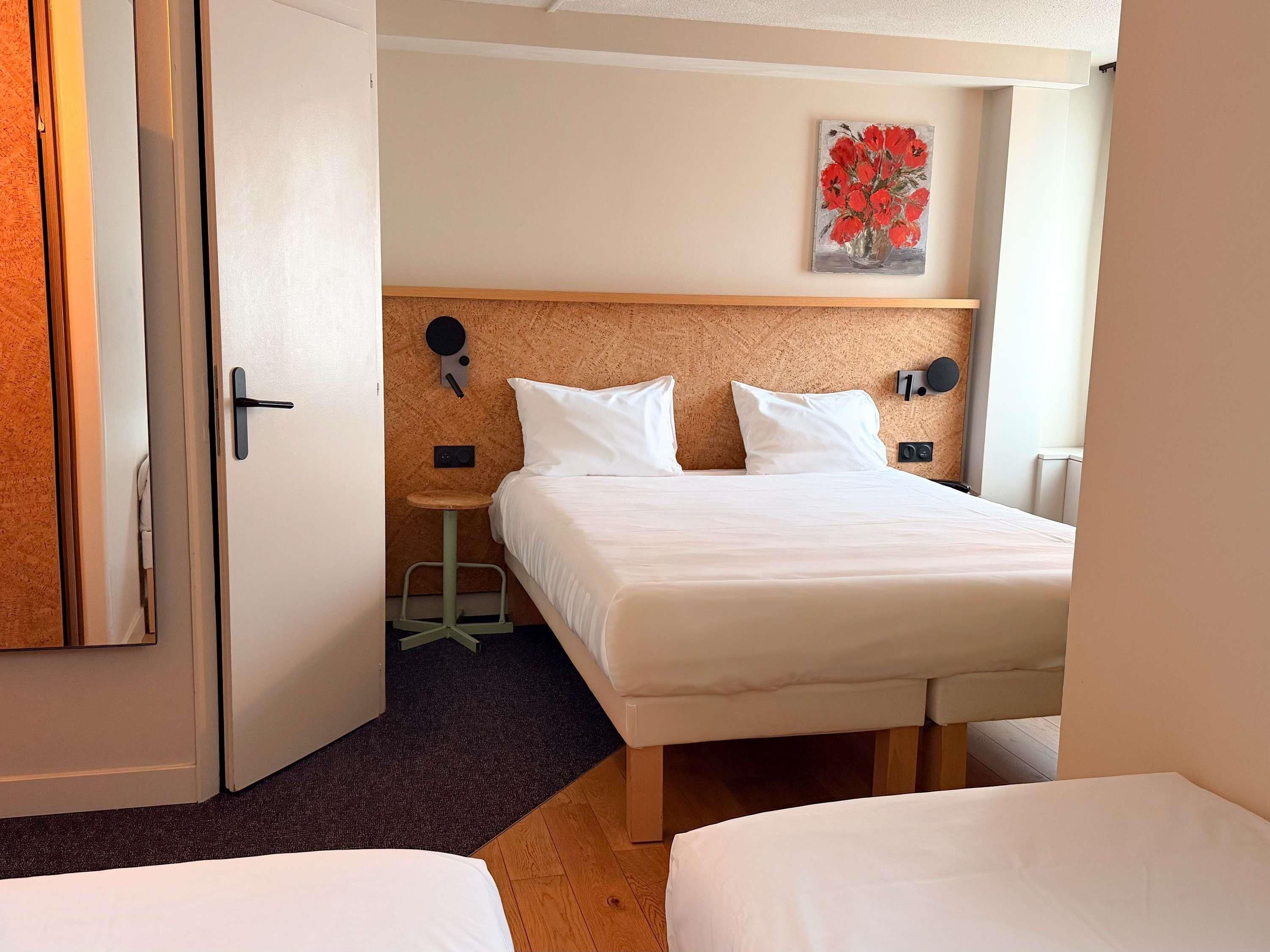Quarto de hotel com cama, uma mesinha, uma cadeira, um relógio de parede e um quadro.