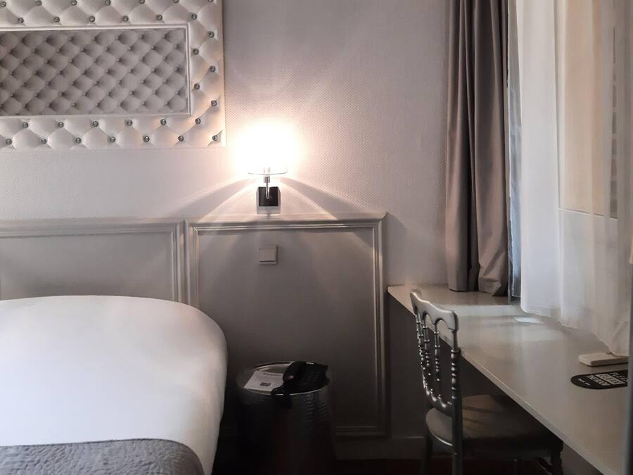 Quarto de hotel com cama, escrivaninha, cadeira, lixeira e luminária fixada na parede.