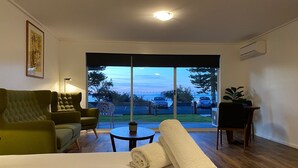 Quarto de hotel com uma cama, duas poltronas, uma mesa pequena, uma planta em vaso e uma janela grande com vista para carros estacionados e árvores.