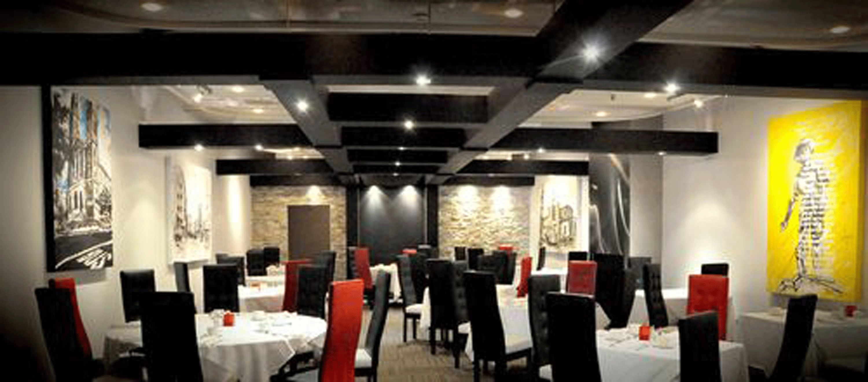 Um restaurante com mesas redondas, cadeiras pretas e vermelhas, e obras de arte nas paredes.