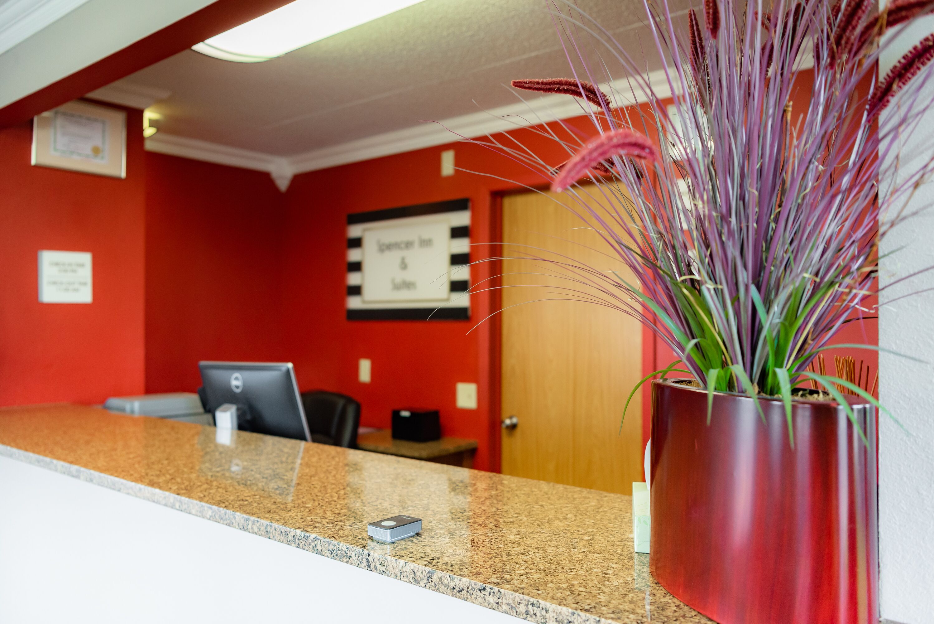 Bancada de recepção com parede vermelha, uma placa emoldurada, um vaso de planta e um computador sobre a mesa.
