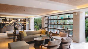 Uma sala de estar moderna com sofá, poltronas e mesa de centro. Há uma estante com objetos decorativos e vista para o verde lá fora.