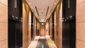 Corredor moderno de hotel com elevadores indicando dos andares 1 ao 16, luminária central e um vaso de planta.
