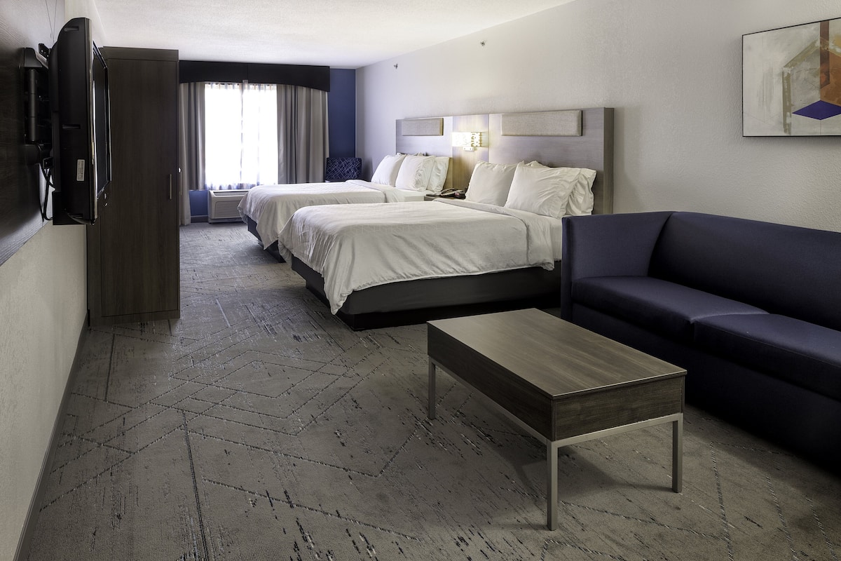 Quarto de hotel com duas camas, um sofá, uma televisão e uma mesa de centro.