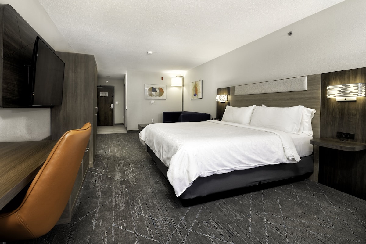 Quarto de hotel moderno com uma cama grande, uma escrivaninha com cadeira, uma televisão de tela plana e um aparelho de luz fixado na parede.