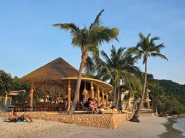 Um resort à beira-mar com um gazebo de telhado de palha, palmeiras e uma praia de areia.