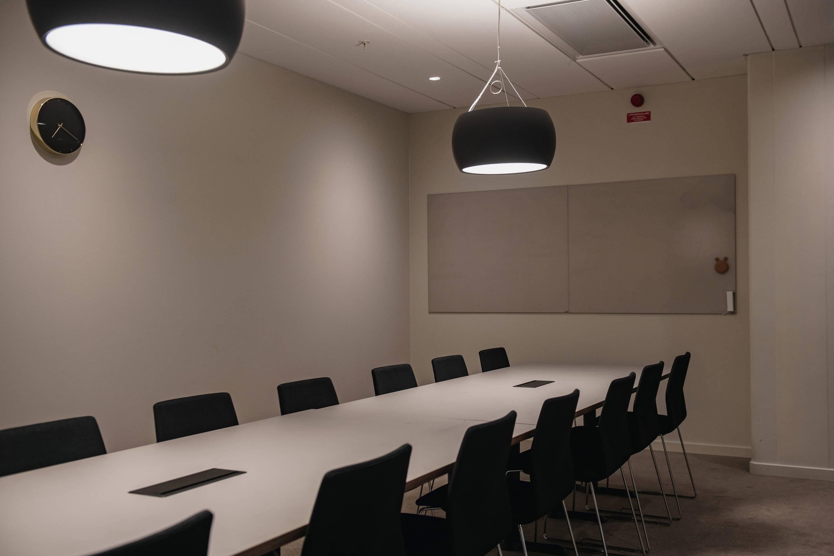 Uma sala de conferências com uma mesa comprida, cadeiras pretas e duas luminárias pendentes.