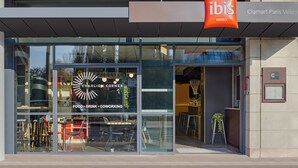 Uma entrada moderna de hotel com fachada de vidro, um letreiro para 'ibis Hotels' e um sinal visível de 'Clamart Paris Vélizy'. No interior, há mesas e cadeiras, indicando uma área de refeições.