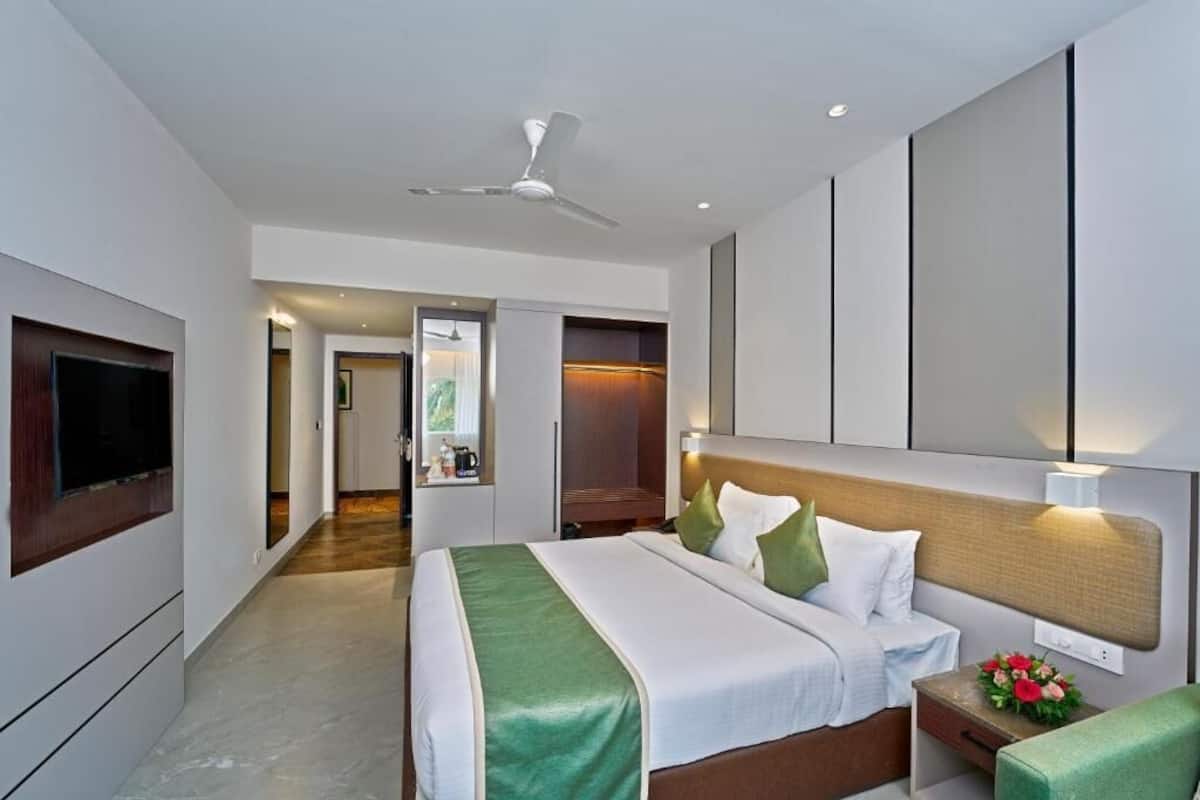 Quarto de hotel com cama, televisão, ventoinha no teto e mesa de cabeceira com arranjo de flores.