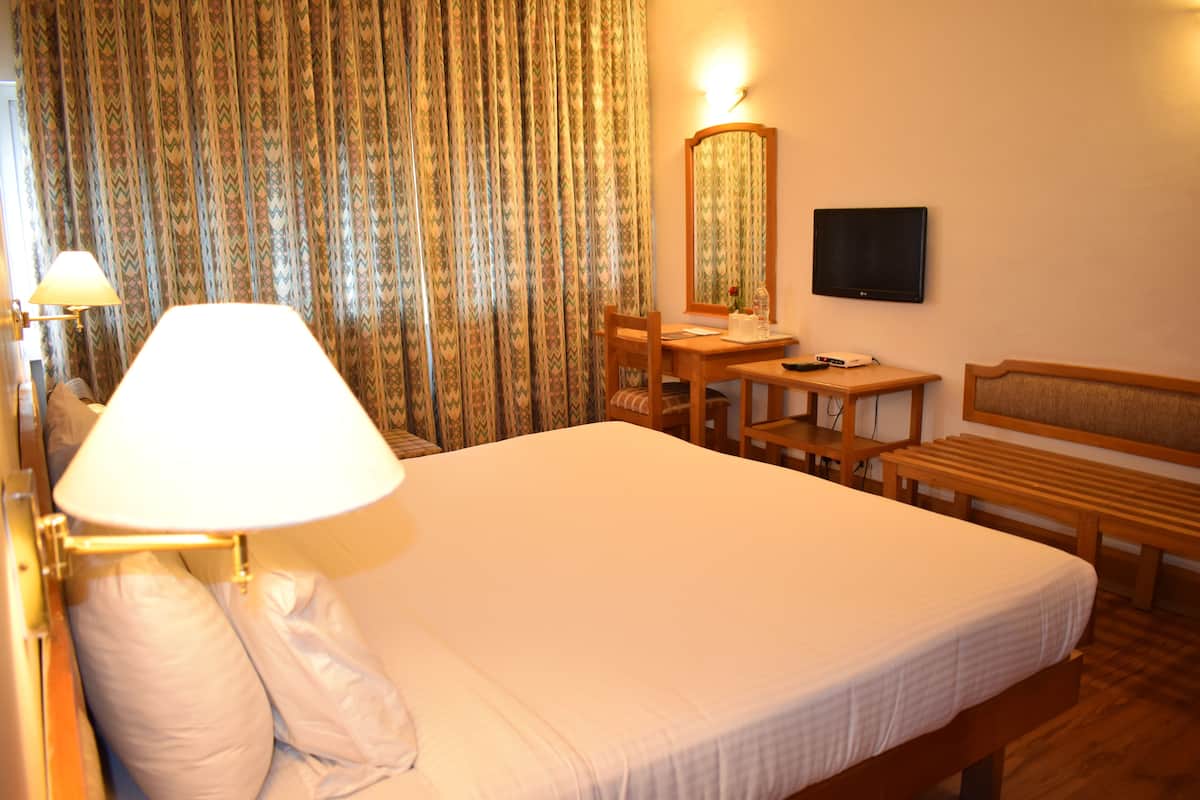 Quarto de hotel com uma cama grande, uma escrivaninha com cadeira, um banco de madeira, uma televisão e cortinas.