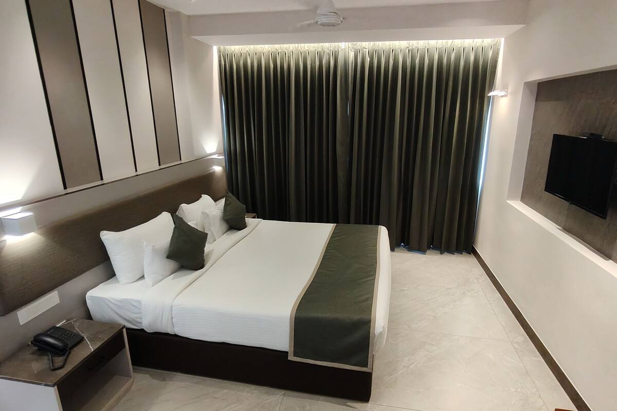 Quarto de hotel moderno com uma cama grande, mesa de cabeceira com telefone, televisão na parede e uma janela com cortinas.