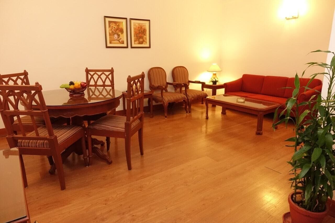 Uma sala de estar com uma mesa de jantar de madeira, seis cadeiras, um sofá vermelho, uma mesa lateral com um abajur e duas fotografias emolduradas na parede.