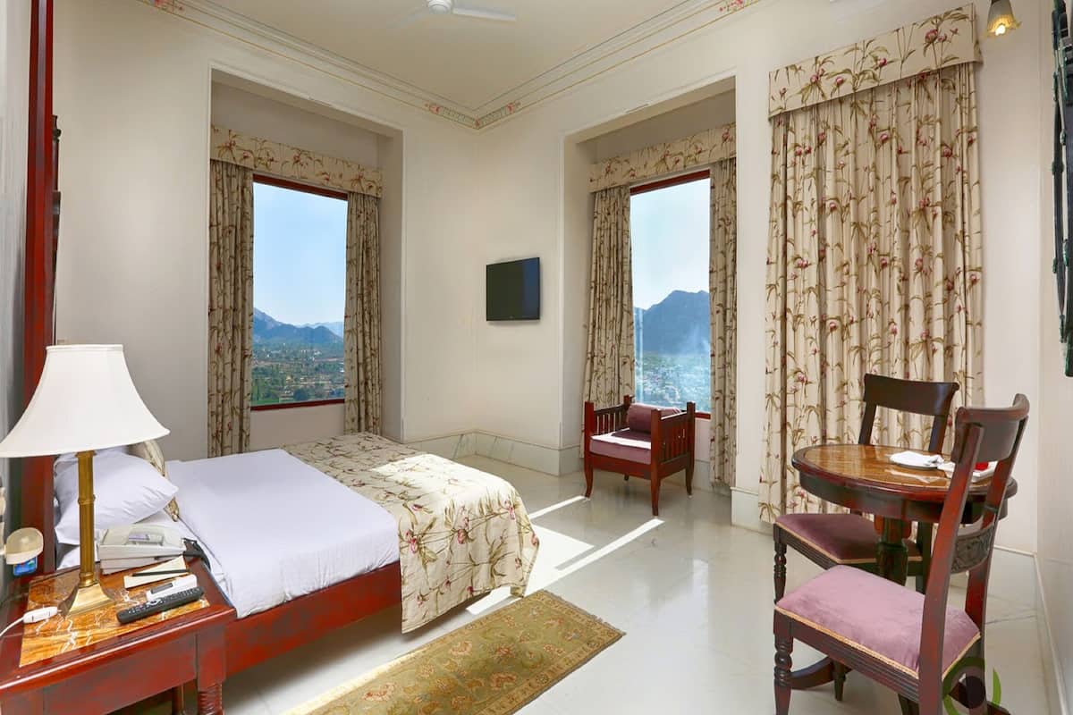 Quarto de hotel com cama, escrivaninha com luminária, cadeira, mesinha e TV. Há duas janelas com cortinas, e é visível uma vista para montanhas e uma cidade através delas.