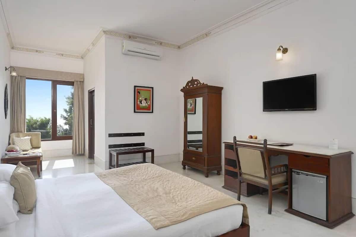 Quarto de hotel com cama, escrivaninha, cadeira, televisão, guarda-roupa e janela.