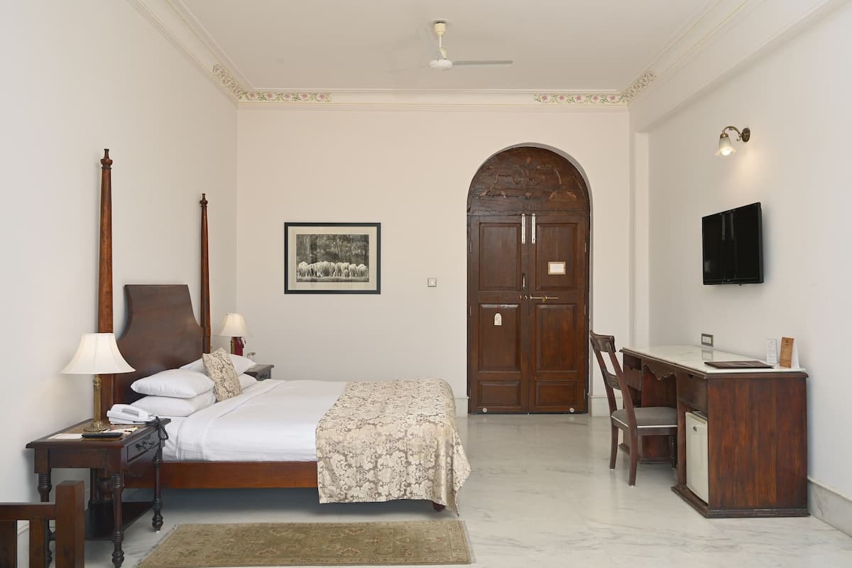 Quarto com cama, mesa de cabeceira, abajur, escrivaninha, cadeira, televisão e porta que dá para outro espaço.