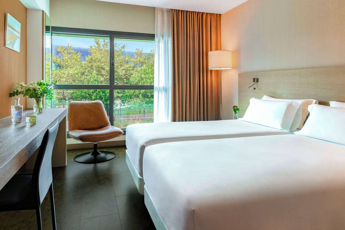 Quarto de hotel com duas camas, uma escrivaninha, uma cadeira, uma janela com cortinas e um abajur.