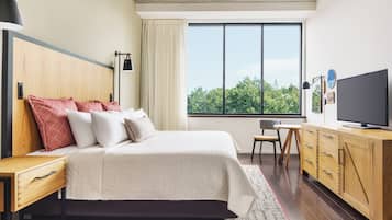 Quarto de hotel moderno com uma cama grande, televisão, escrivaninha e vista para o verde lá fora.
