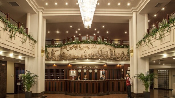 Um hall espaçoso com um lustre imponente, um balcão de recepção e teto alto com molduras decorativas.
