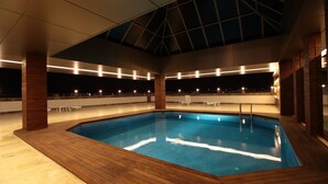 Piscina interior com deck de madeira, teto de vidro e vista noturna da cidade.