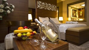 Quarto de hotel com mesa de madeira, tigela de frutas, garrafa de vinho e cama com cabeceira.