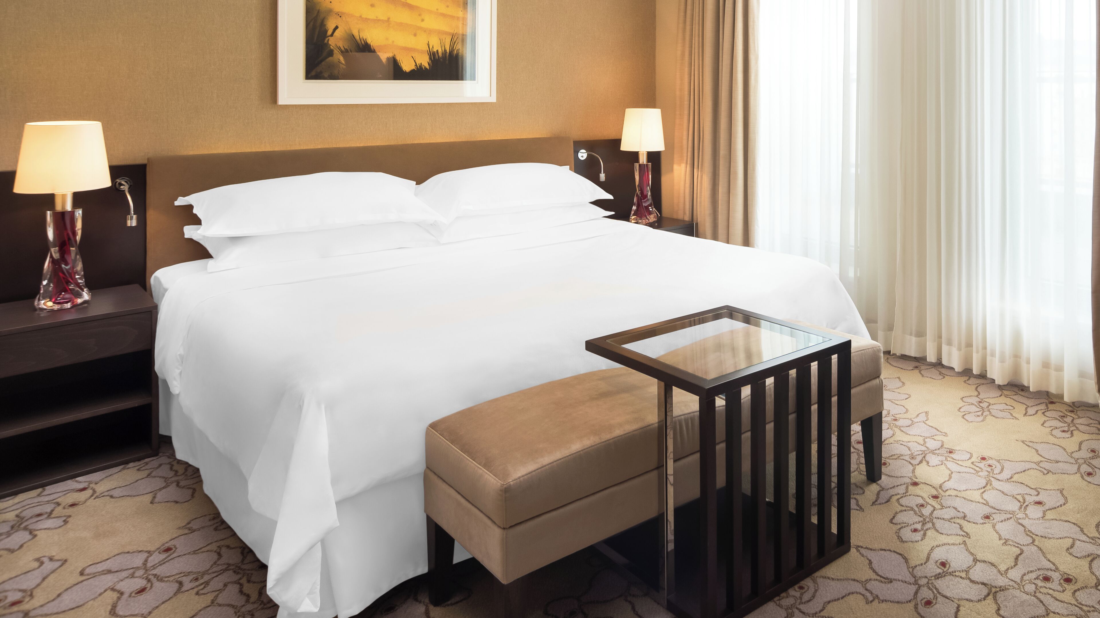 Quarto de hotel com uma cama grande, duas luminárias de cabeceira, uma mesa de vidro, um banco e um quadro na parede.