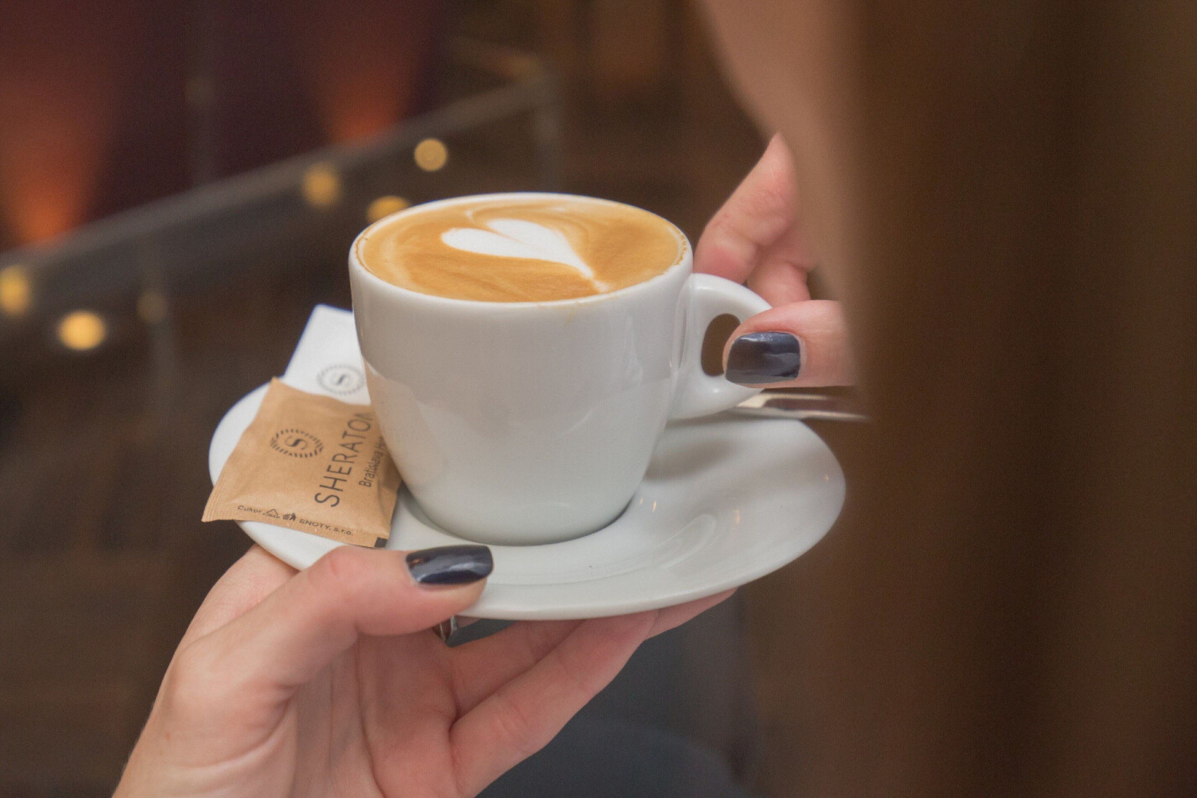 Uma mão segurando uma xícara de café com arte de latte em formato de coração e um sachê de café Sheraton.