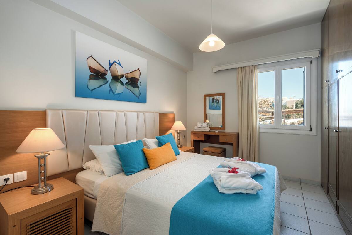 Quarto de hotel com cama, mesas de cabeceira, luminária, pintura, escrivaninha, cadeira, janela e guarda-roupa.