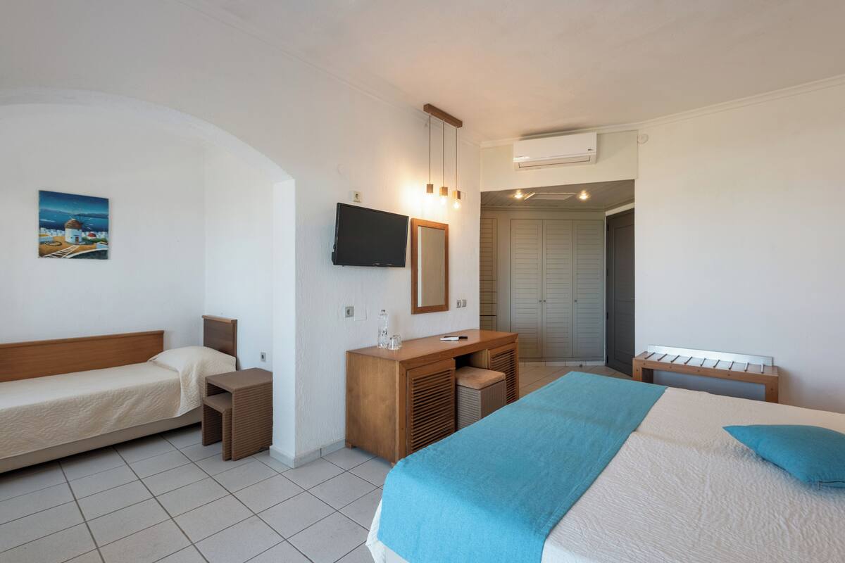 Quarto de hotel com cama, escrivaninha, cadeira, televisão e banheiro visível através de uma porta em arco.