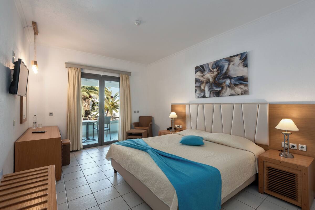 Quarto de hotel com cama, escrivaninha, cadeira, televisão e varanda com vista para palmeiras.