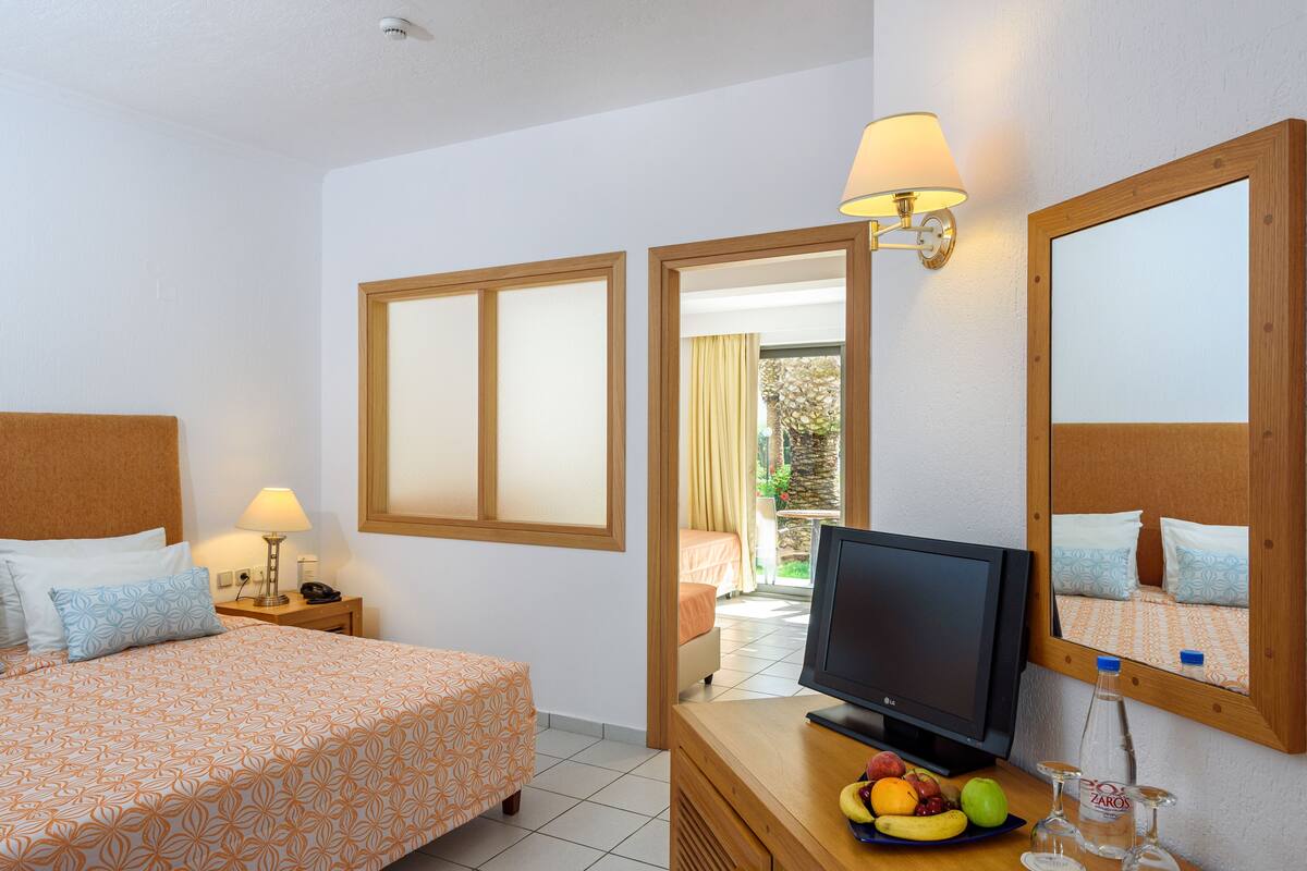 Quarto de hotel com cama, televisão, mesa de cabeceira com abajur e uma cesta de frutas.