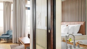 Quarto de hotel com um banco de madeira, uma escrivaninha, uma cadeira, uma cama com cabeceira e um banheiro com pia e espelho.