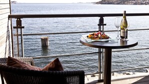 Uma varanda com uma cadeira de vime, uma mesa com garrafa de vinho, copos e frutas.