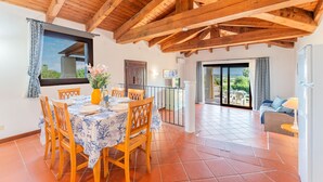 Área de refeições com uma mesa posta para quatro pessoas, cadeiras de madeira e vista pela janela.