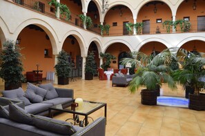 Amplo espaço interno com vários arcos, área de estar com sofás e uma mesa de centro, e área de piscina com plantas de palmeira.