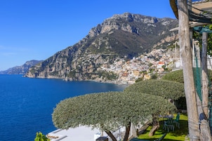 Uma vila costeira com uma montanha imponente ao fundo, um mar azul límpido e uma área de jardim bem cuidada com uma mesa e cadeiras.