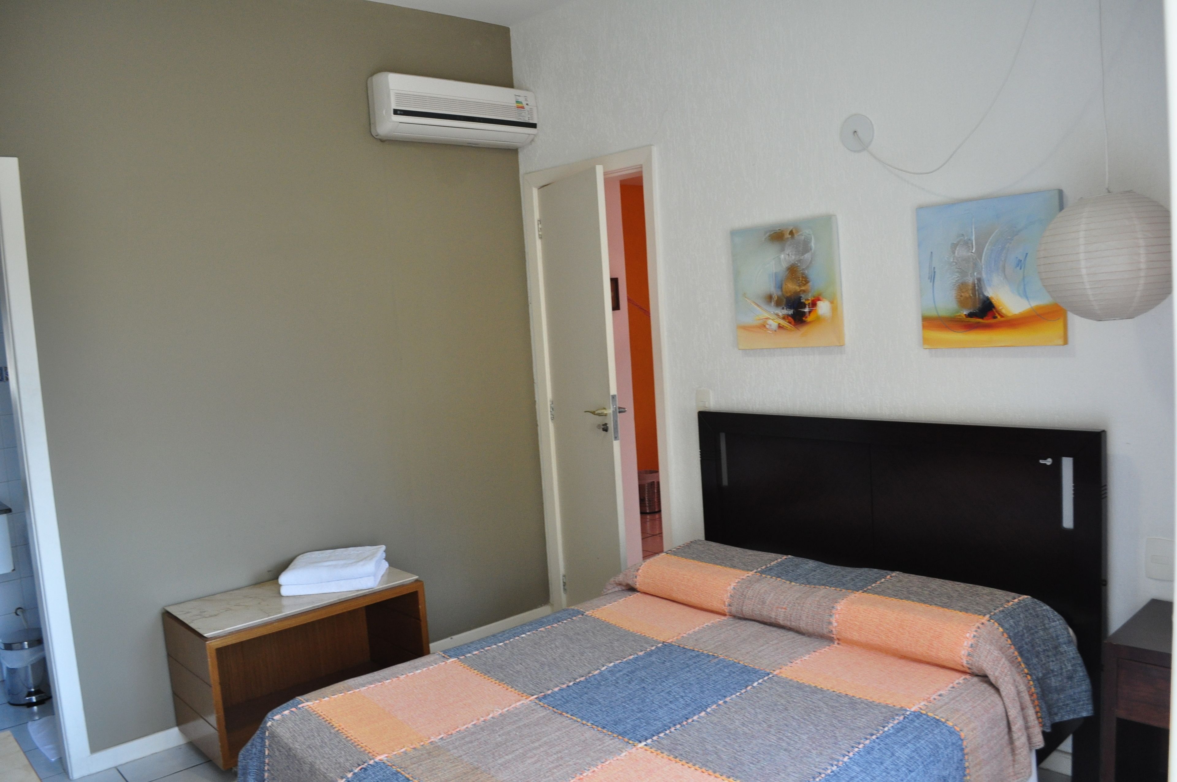 Quarto com cama, mesa de cabeceira com toalha, aparelho de ar condicionado e duas pinturas na parede.