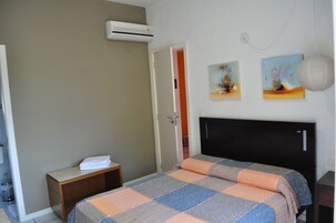 Quarto com cama, mesa de cabeceira com toalha, aparelho de ar condicionado e duas pinturas na parede.