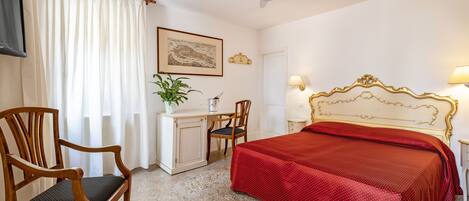 Quarto de hotel com cama, escrivaninha, cadeira, ventilador de teto e janela com cortinas.