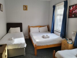 Quarto de hotel com duas camas de solteiro, uma cabeceira de madeira, uma mesinha de cabeceira com um tabuleiro de chávenas brancas e uma janela com cortinas azuis.