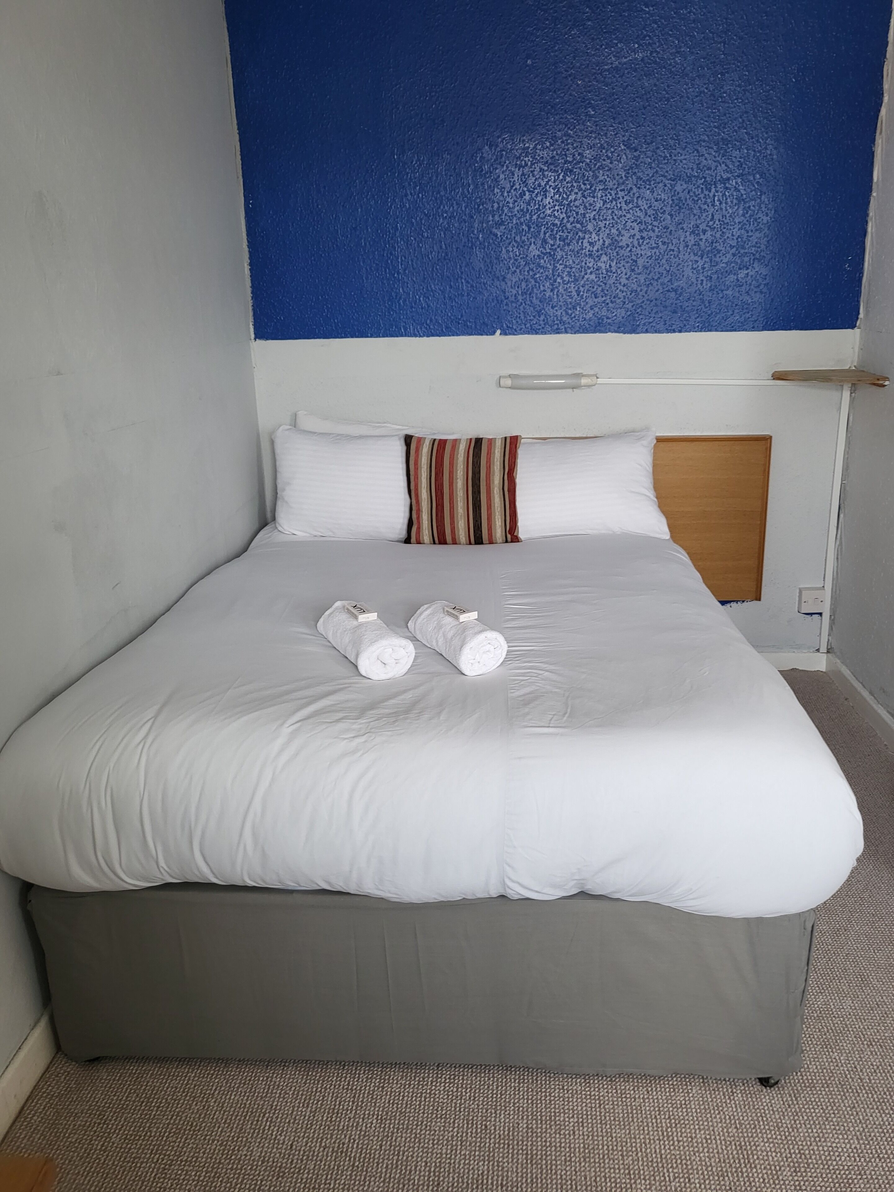 Cama bem arrumada com lençóis brancos e duas toalhas enroladas, cabeceira de madeira e parede azul.