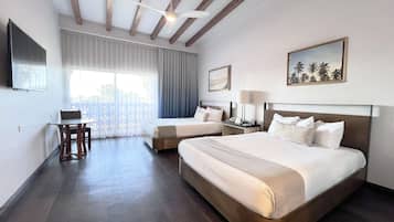 Quarto de hotel com duas camas, uma escrivaninha, uma cadeira, um ventilador de teto e uma janela grande com cortinas.