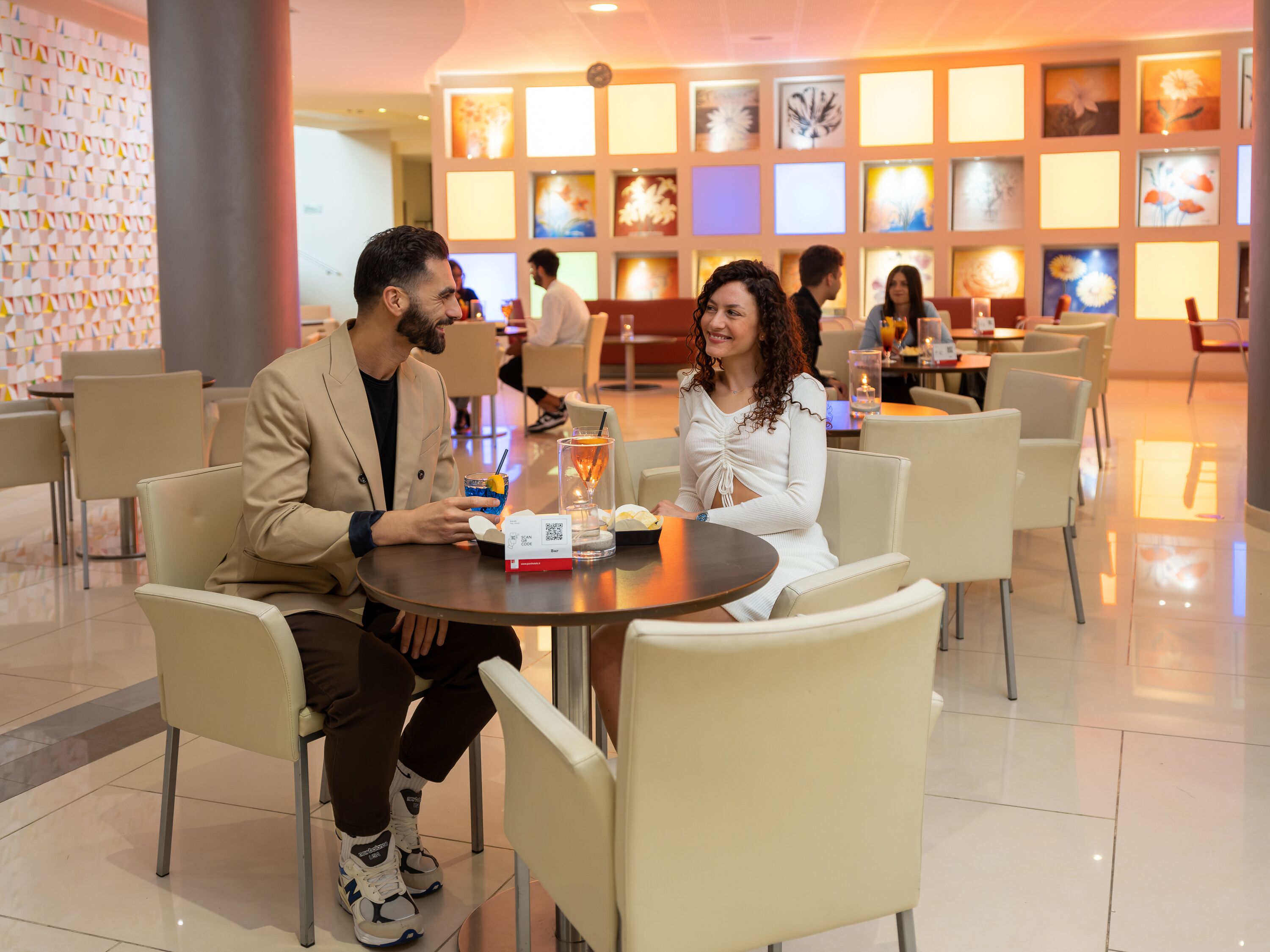 Um café moderno com um design limpo e minimalista, apresentando uma mesa redonda central com cadeiras, uma parede decorativa com obras de arte emolduradas e um chão polido.