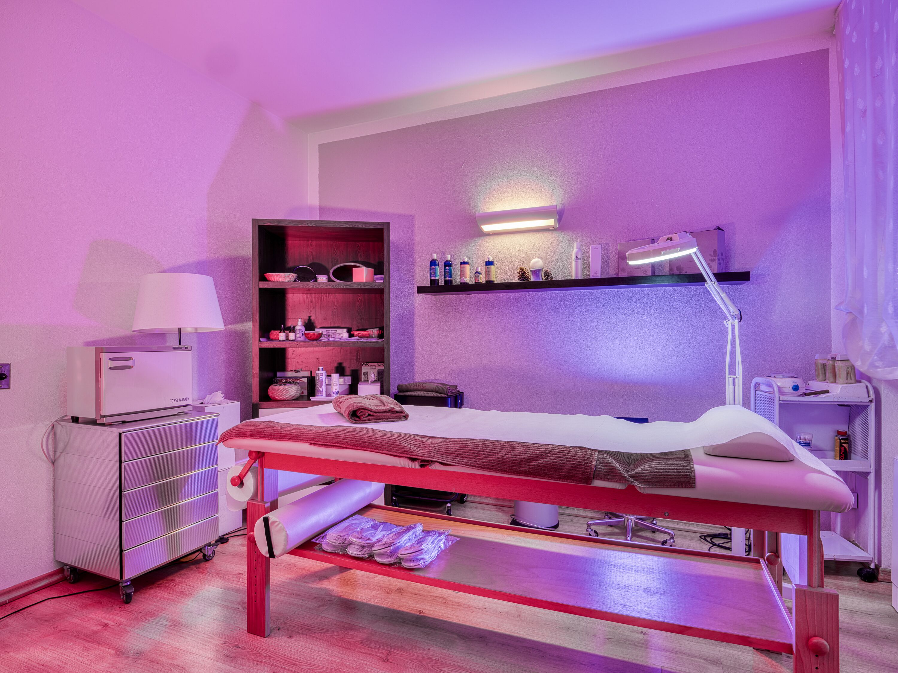 Uma sala de exames médicos com uma cama, mesa de exame, prateleiras com materiais médicos e um armário com gavetas.