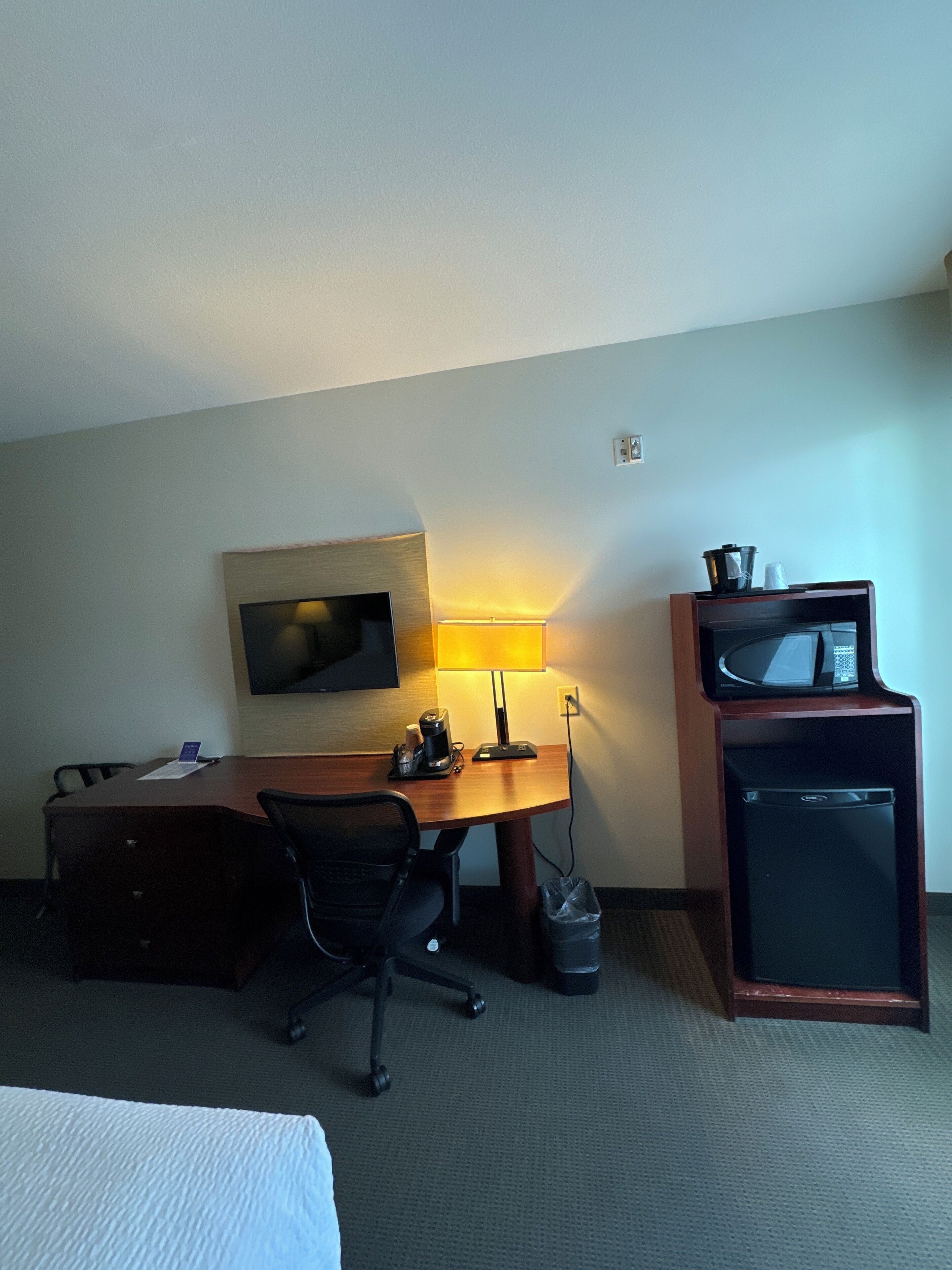 Quarto de hotel com uma escrivaninha de madeira, uma cadeira de escritório preta, uma televisão na parede, um pequeno frigorífico e um micro-ondas.