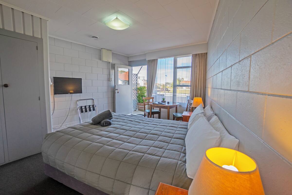 Quarto de hotel com cama, televisão, mesa pequena e cadeiras, e janela com cortinas.