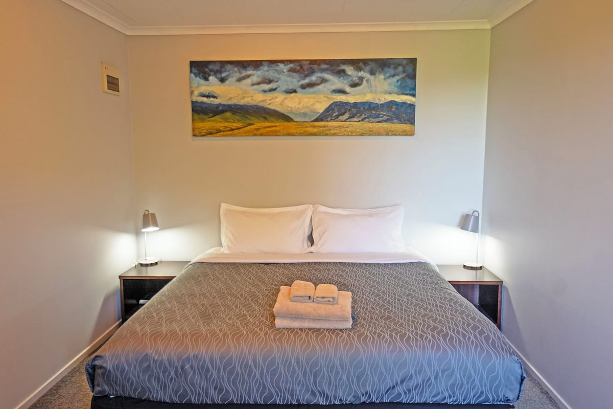 Quarto de hotel com uma cama, duas luminárias de cabeceira, um quadro na parede e uma toalha dobrada sobre a cama.