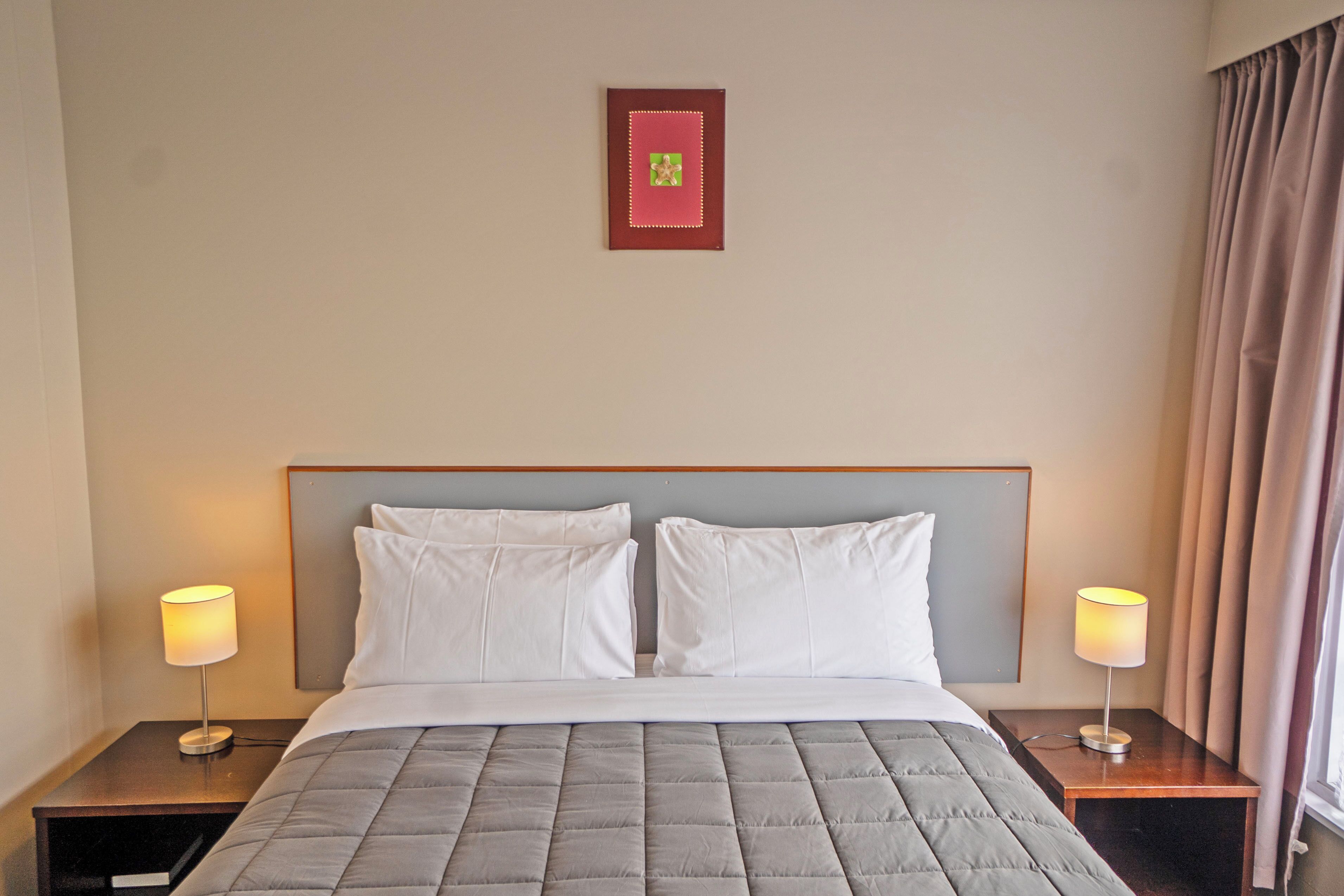 Cama bem arrumada com um edredom cinza, dois travesseiros brancos e duas luminárias de cabeceira sobre mesas de madeira.
