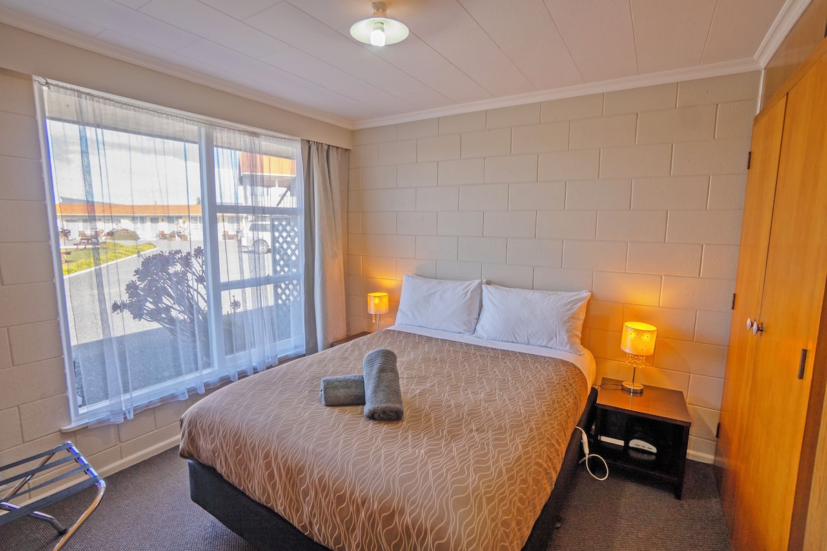 Quarto de hotel com uma cama grande, duas luminárias de cabeceira, um guarda-roupa de madeira e uma janela com cortinas.